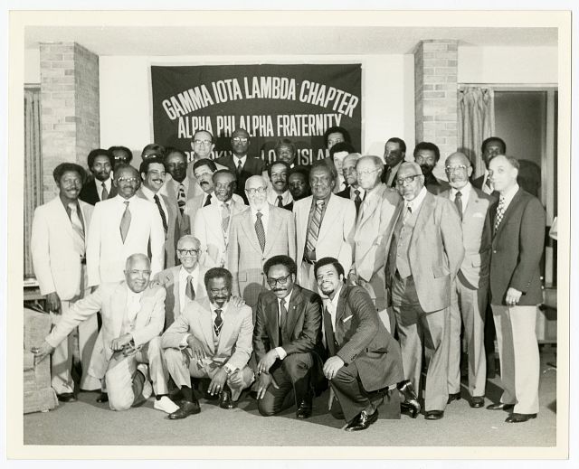 Large group of men pose for a portrait in front of a banner reading ...